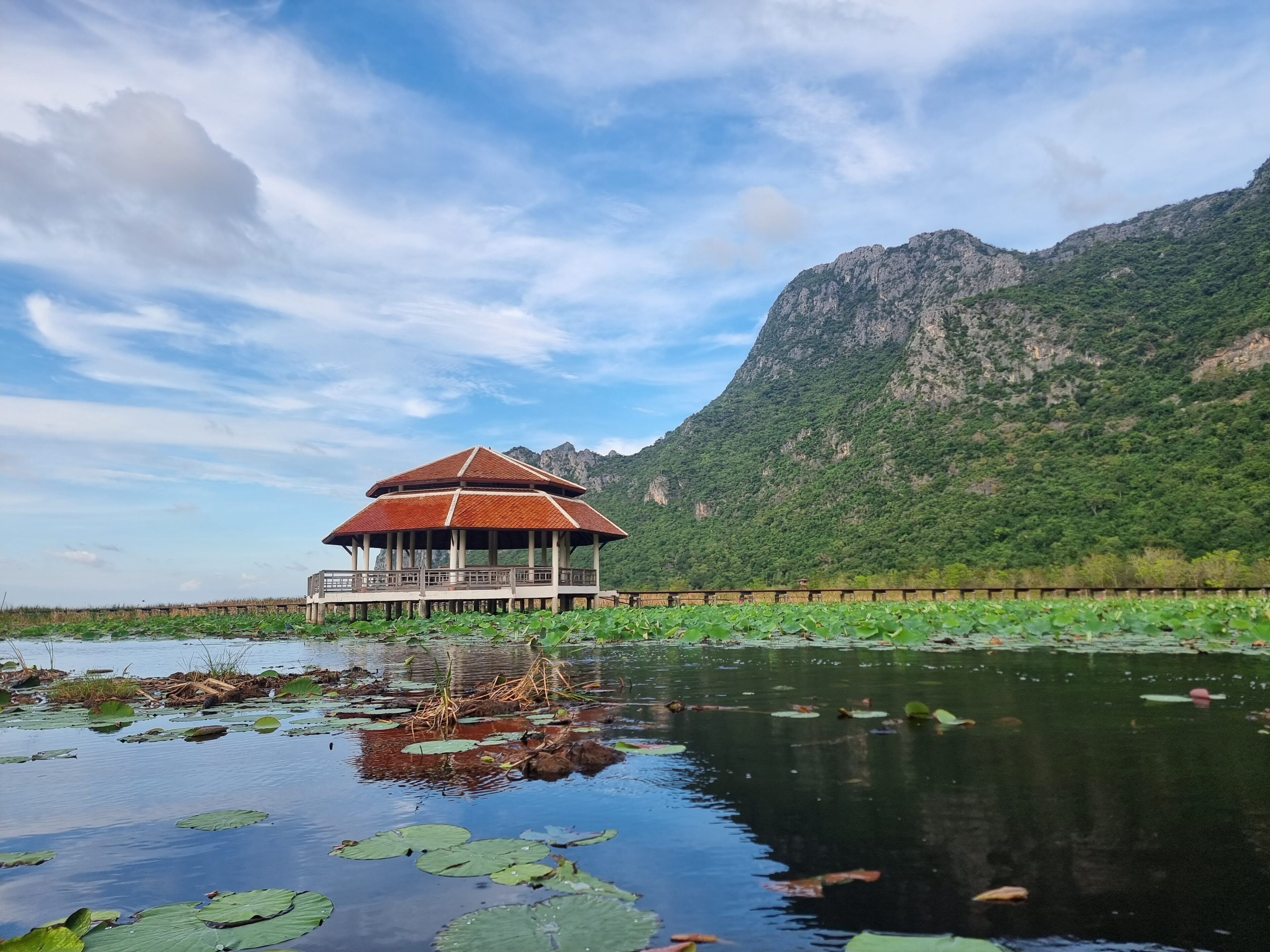 Um pavilhão com telhado vermelho e paredes brancas, rodeado por água e vegetação, com uma montanha ao fundo.