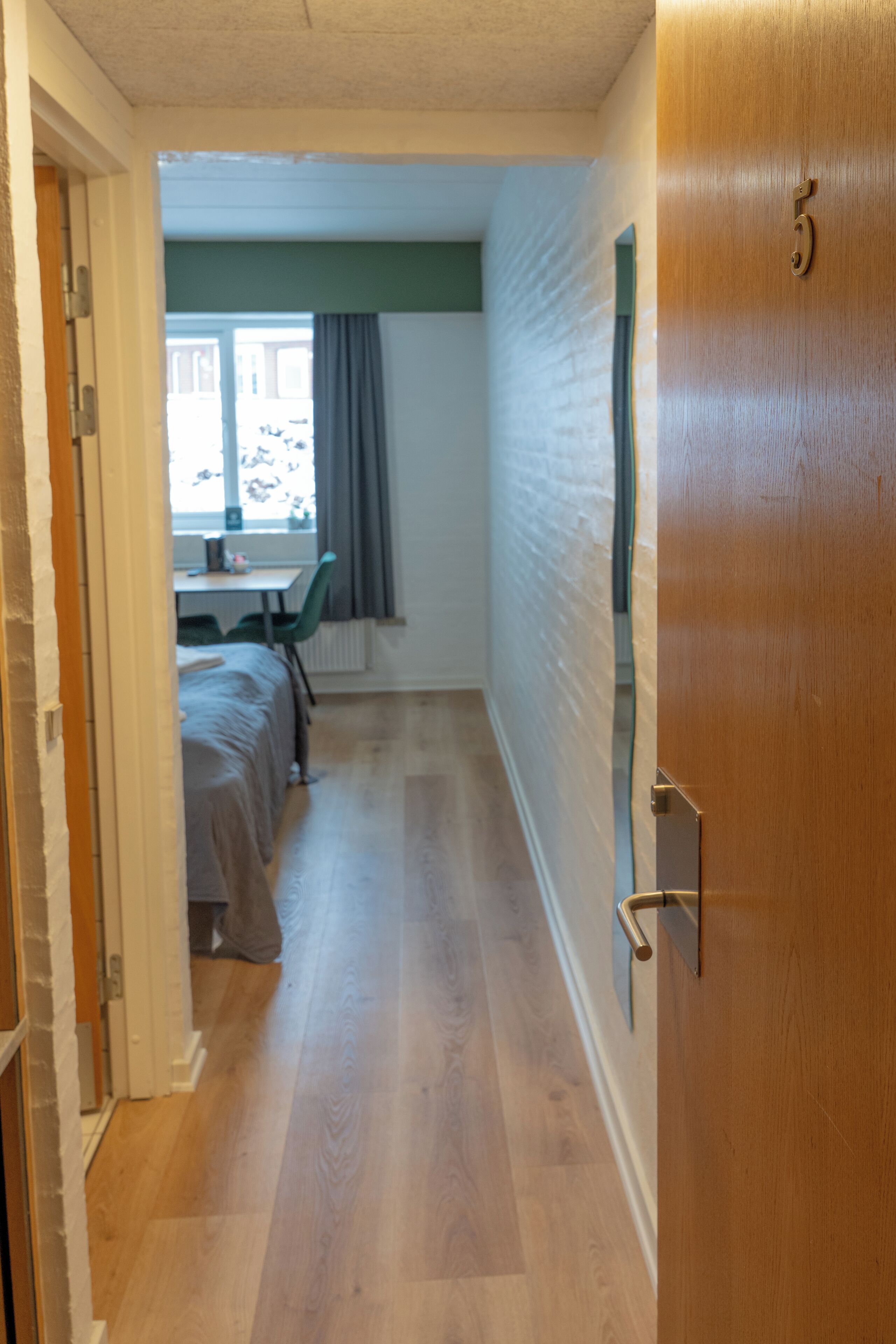 Um corredor estreito com uma porta de madeira à direita, uma cama com colcha cinza à esquerda e uma janela com cortinas ao fundo.