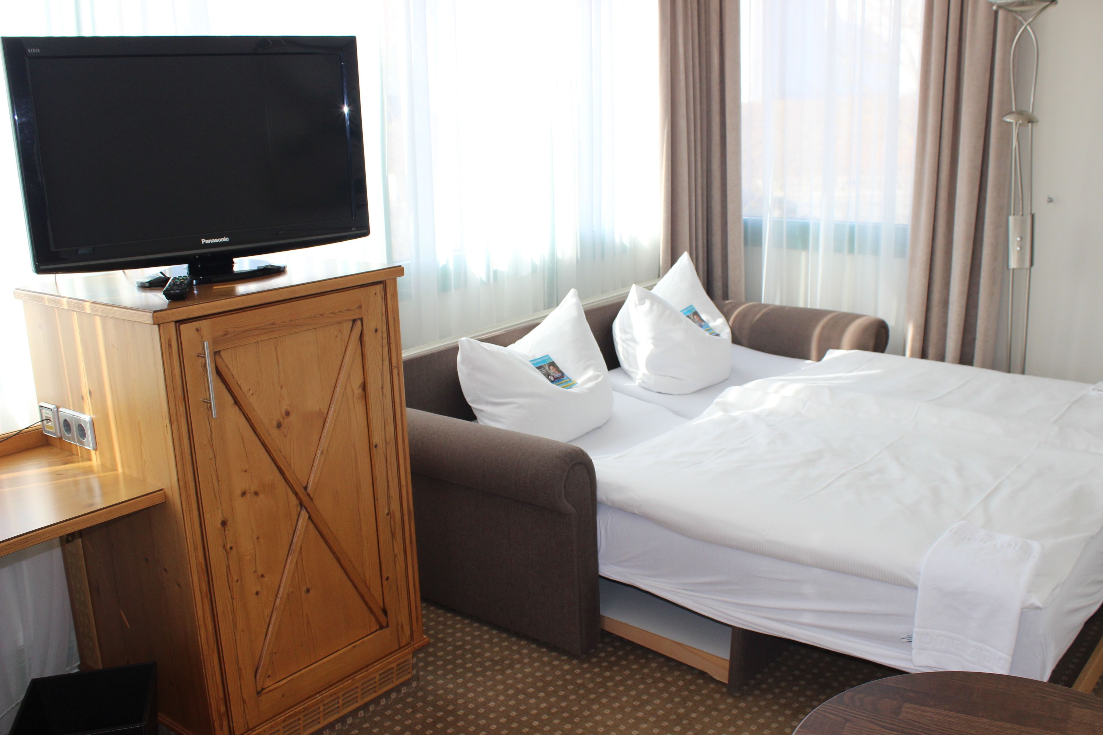 Quarto de hotel com uma cama, uma televisão em um móvel de madeira e uma janela com cortinas.