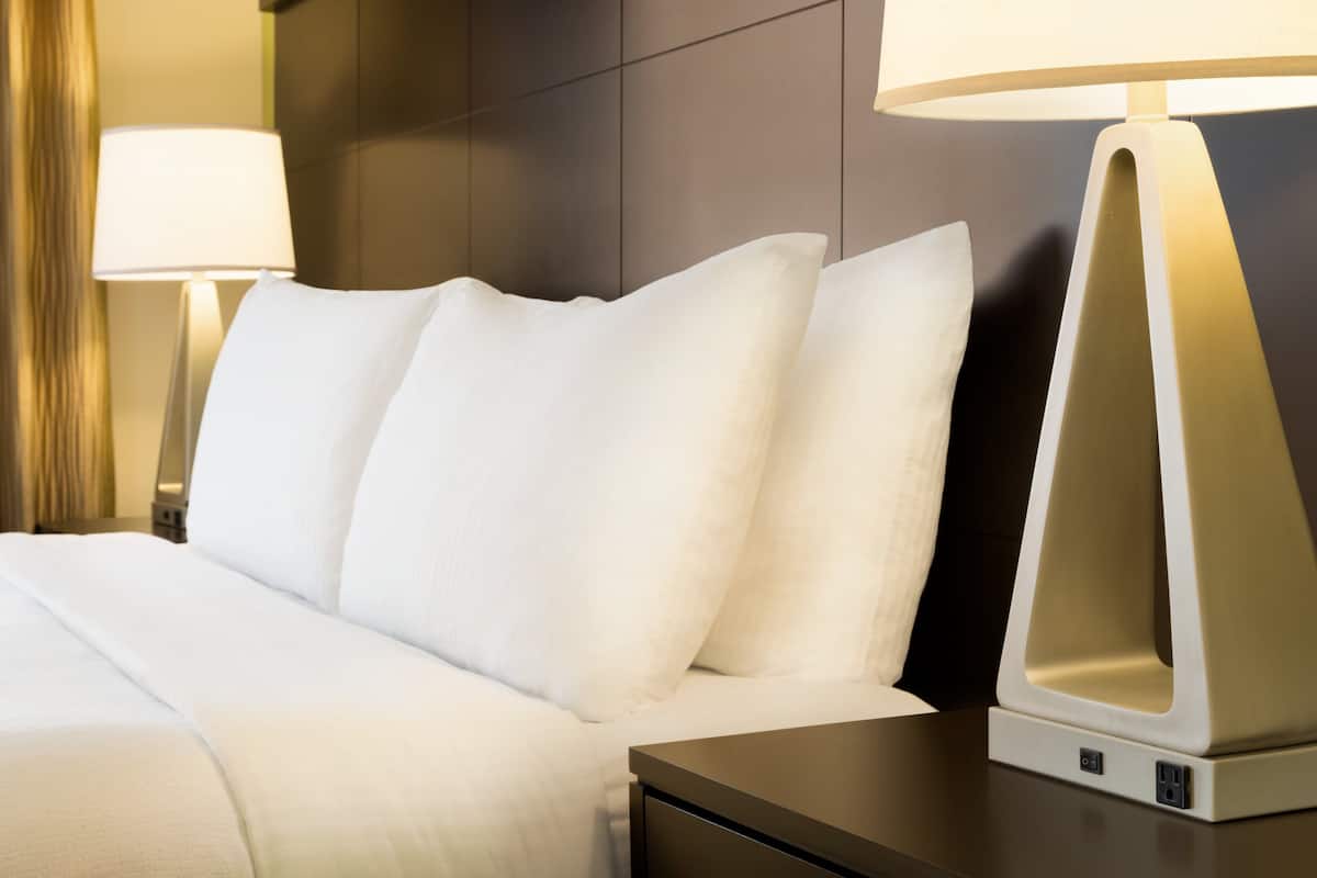 Cama bem arrumada com lençóis brancos, mesa de cabeceira com abajur e luminária na parede.