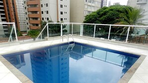 Piscina retangular com grade branca, rodeada por um deck revestido com azulejos.