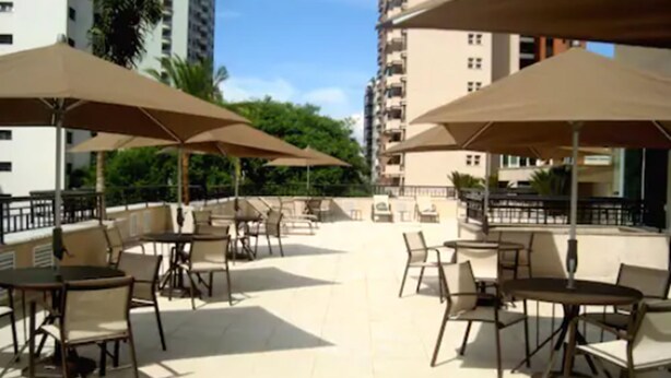 Uma área de terraço ao ar livre com mesas e cadeiras sob guarda-sóis, rodeada por edifícios altos.