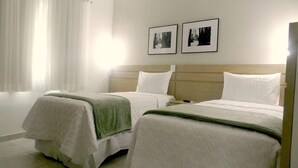 Quarto de hotel com duas camas, uma janela grande com cortinas brancas e duas fotos emolduradas na parede.