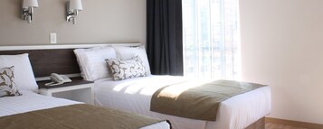 Quarto de hotel com duas camas, mesa de cabeceira com telefone e janela com cortinas.