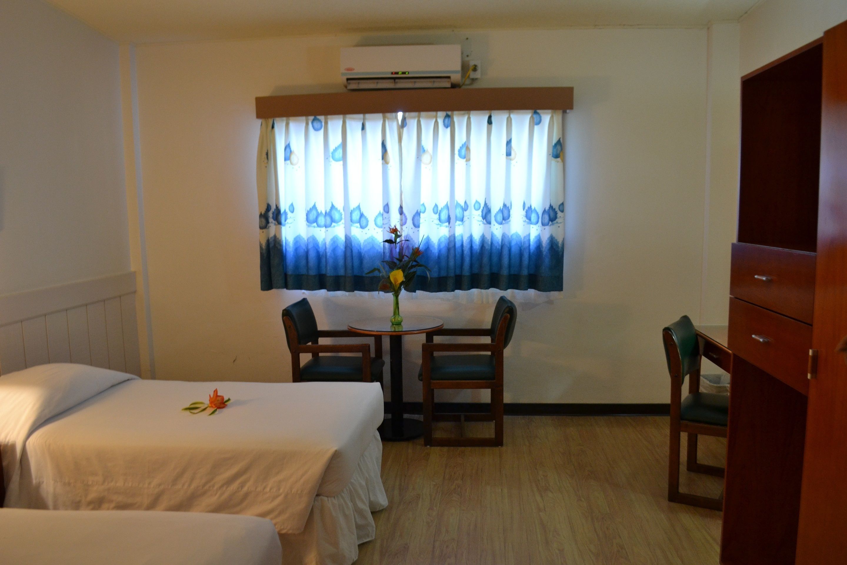 Quarto de hotel com duas camas, uma mesinha com cadeiras, uma janela com cortinas e um aparelho de ar condicionado.