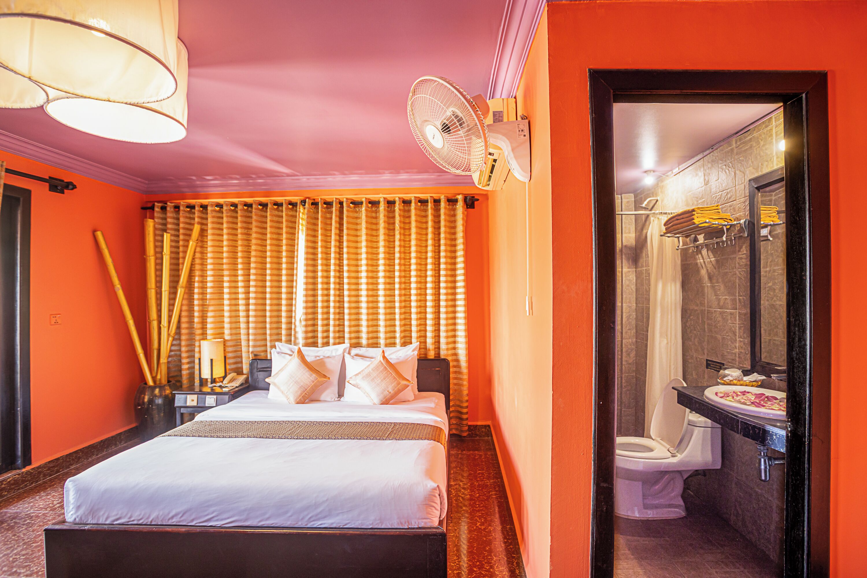 Quarto de hotel com cama, mesa de cabeceira, abajur e um ventilador.
