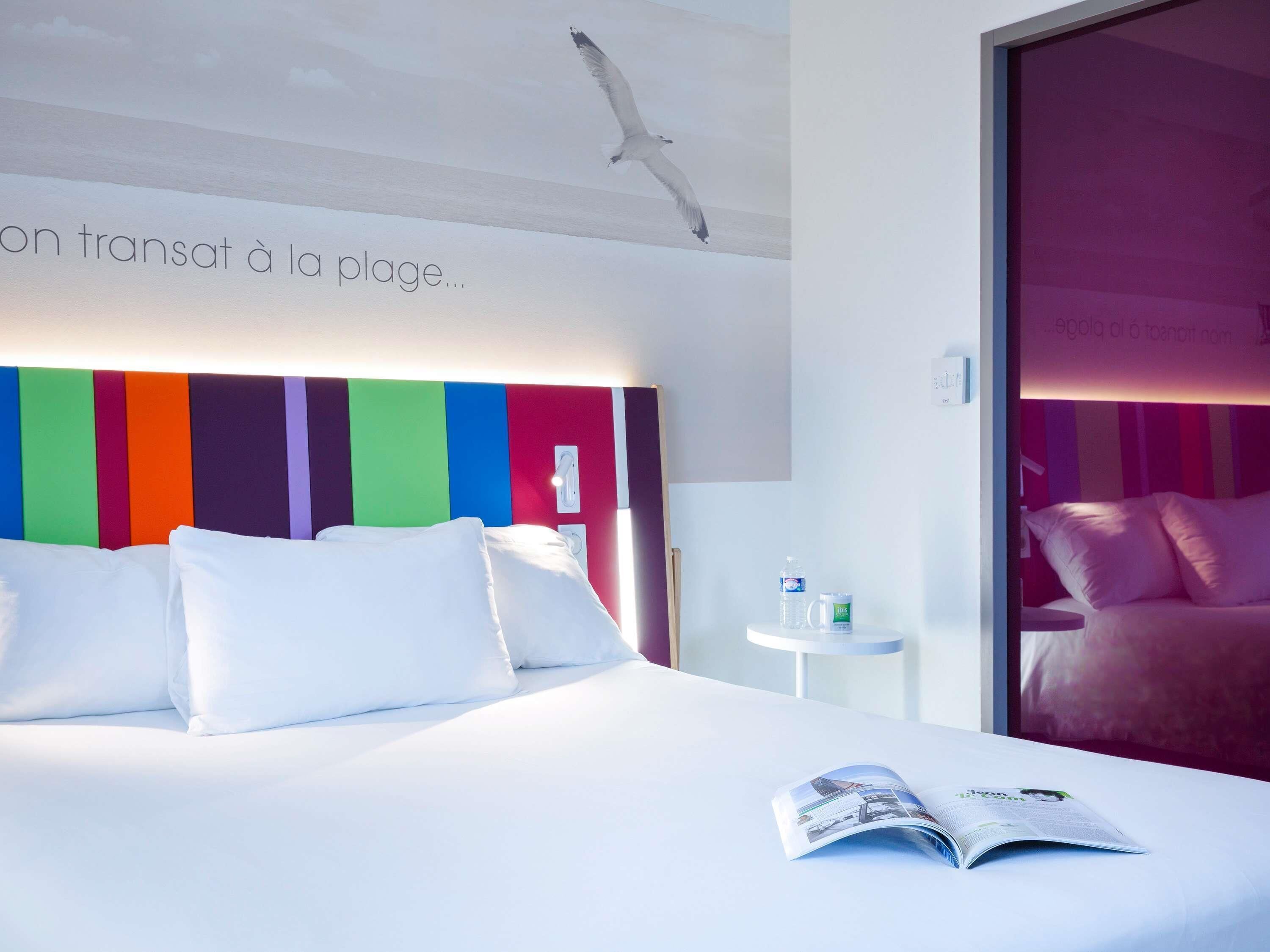 Quarto de hotel com uma cama, uma revista sobre a cama, uma mesinha com uma garrafa e uma chávena, e uma cabeceira listrada e colorida.