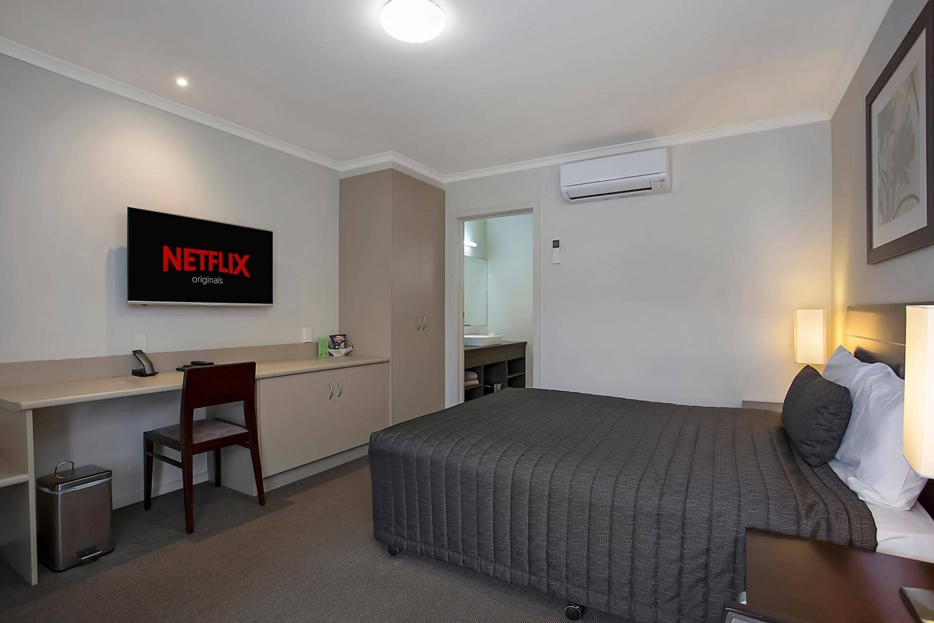 Quarto de hotel com cama, escrivaninha, cadeira e TV exibindo 'NETFLIX originals', além de um ar condicionado.