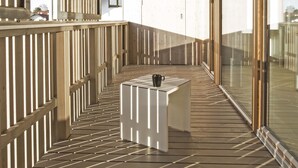 Um deck de madeira com uma mesa e cadeiras brancas, grandes janelas e vista do céu.