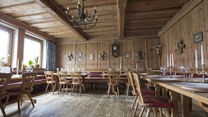 Uma sala de jantar com painéis de madeira, mesas e cadeiras de madeira, um candelabro e relógios fixados na parede.
