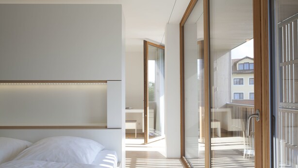 Quarto moderno com uma cama grande, porta de vidro que dá para uma varanda e vista dos edifícios externos.