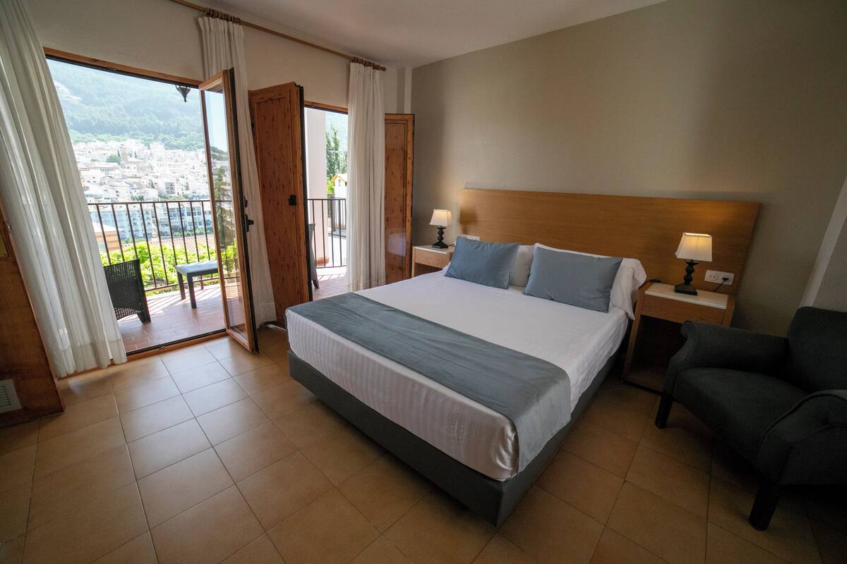 Quarto de hotel com uma cama grande, duas mesas de cabeceira com luminárias, uma cadeira e varanda com vista para a cidade e montanhas.