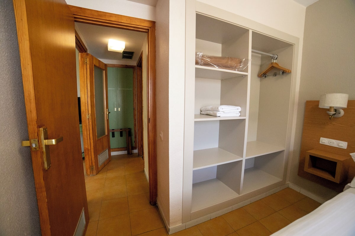 Quarto de hotel com closet espaçoso, uma cama, mesa de cabeceira com abajur e um criado-mudo de madeira.