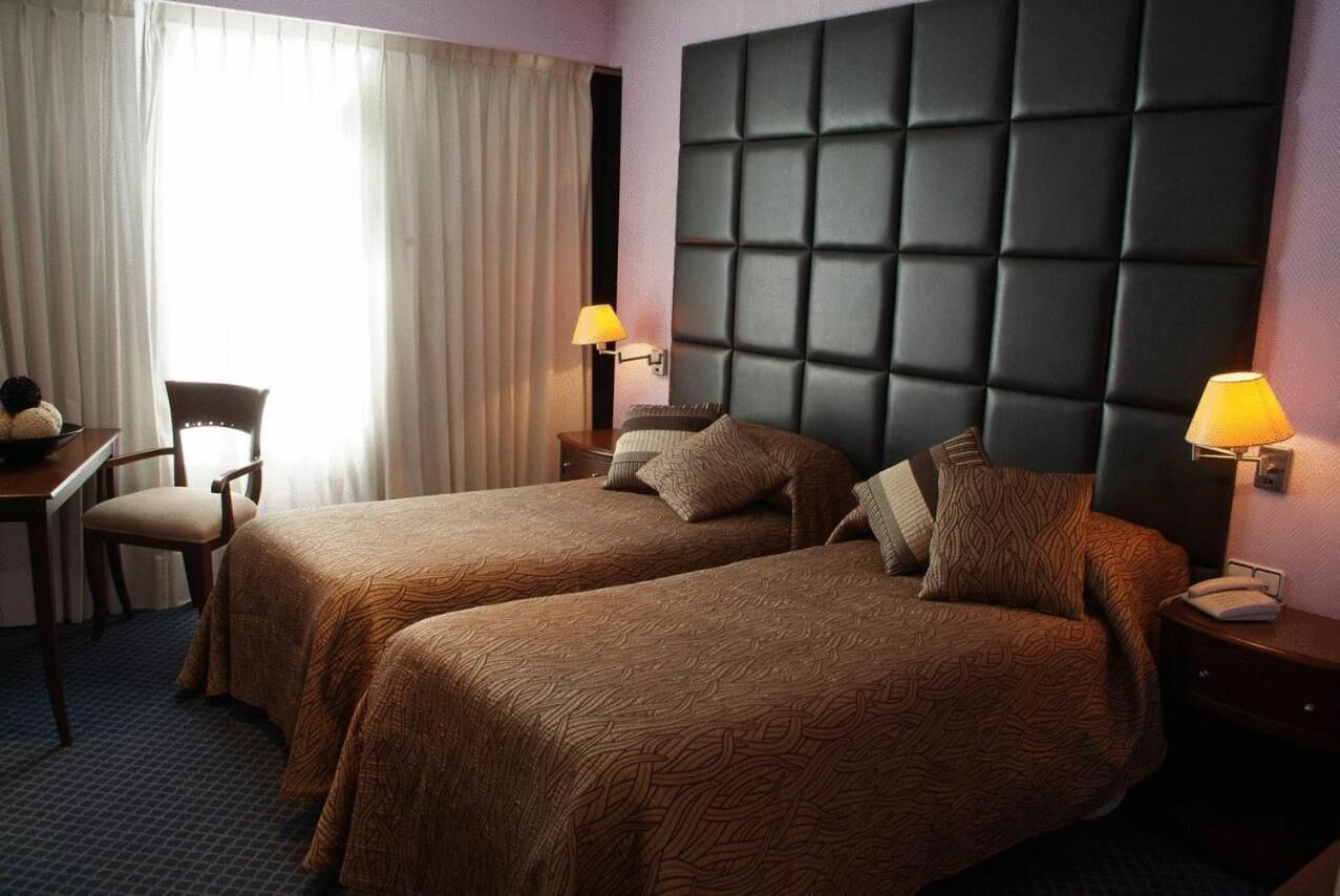 Quarto de hotel com duas camas, uma escrivaninha, cadeiras e luminárias.