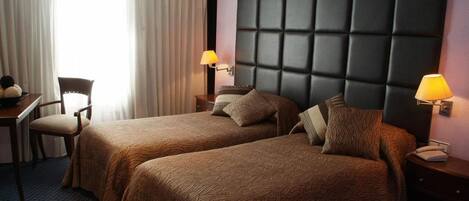 Quarto de hotel com duas camas, uma escrivaninha, cadeiras e luminárias.