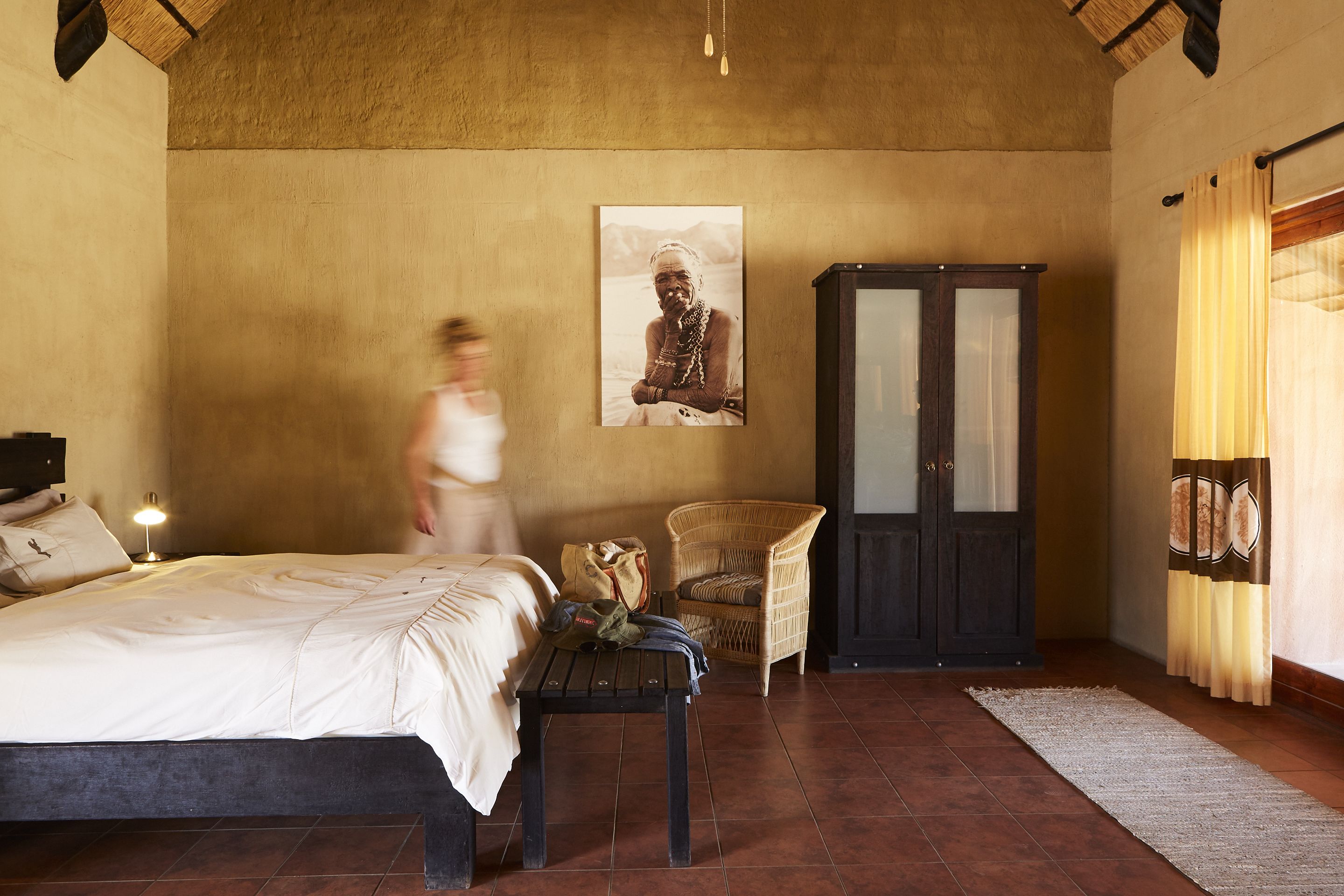 Quarto com cama, um banco de madeira, uma cadeira de vime, guarda-roupa e um cartaz na parede.