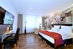 Quarto de hotel com uma cama, uma escrivaninha com cadeira, uma televisão e uma pequena mesa com um vaso.