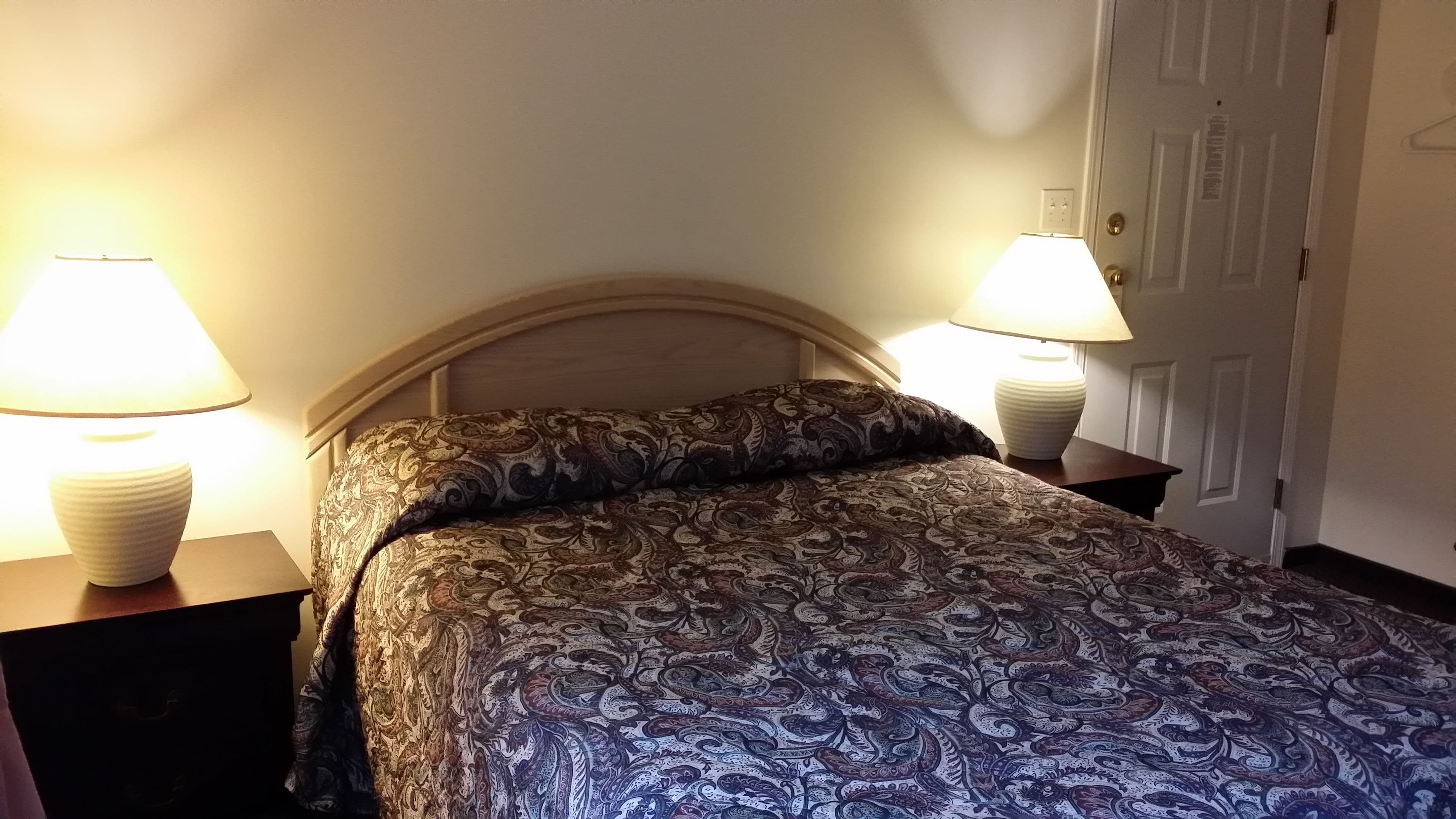 Uma cama com colcha estampada, duas luminárias de cabeceira e uma porta ao fundo.