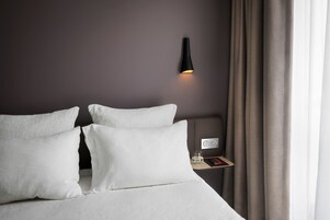 Cama bem arrumada com lençóis e almofadas brancas, mesa de cabeceira com um copo de água e um livro, e luminária moderna fixada na parede.