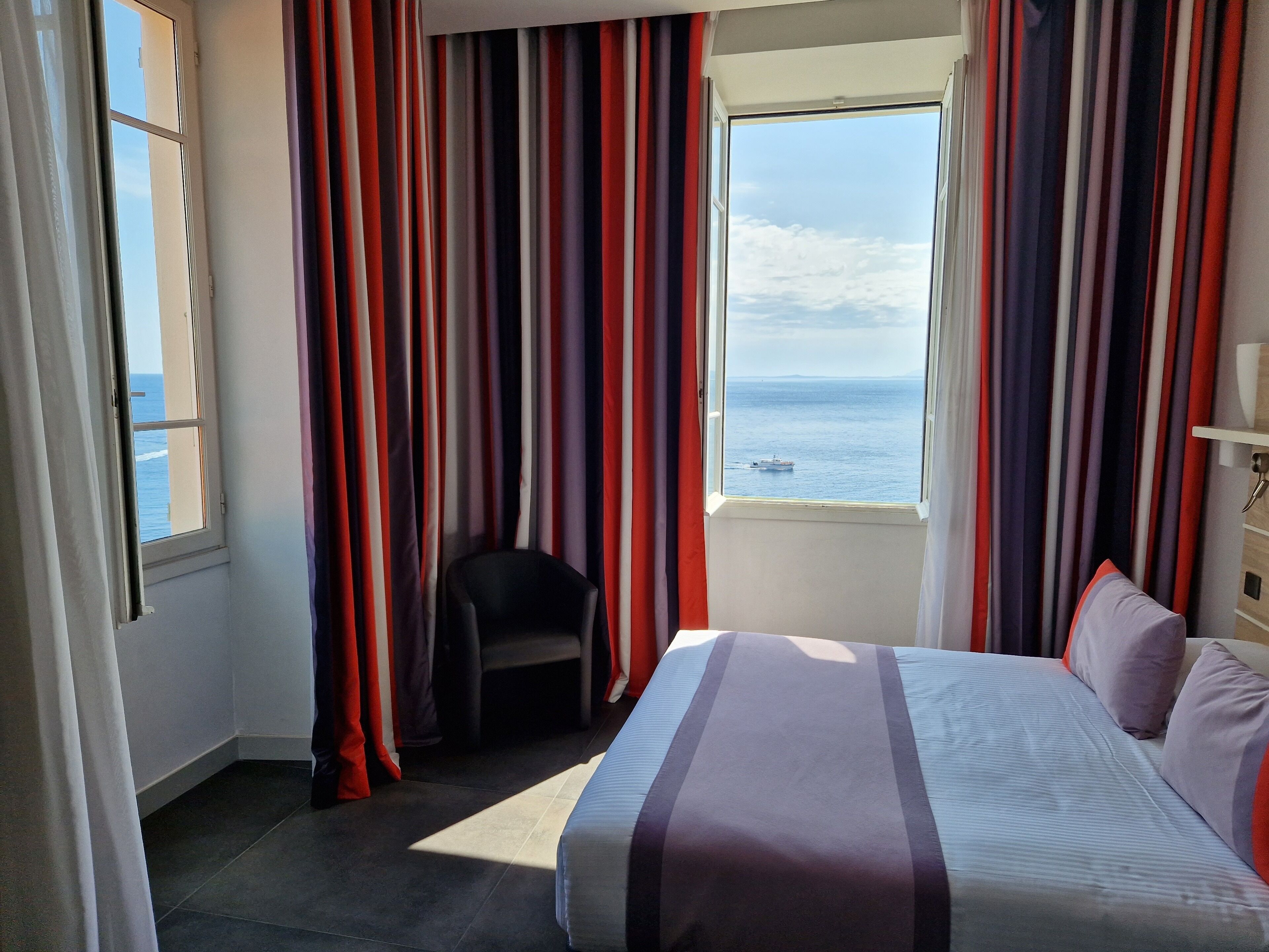 Quarto de hotel com cama, cadeira e janela com cortinas.
