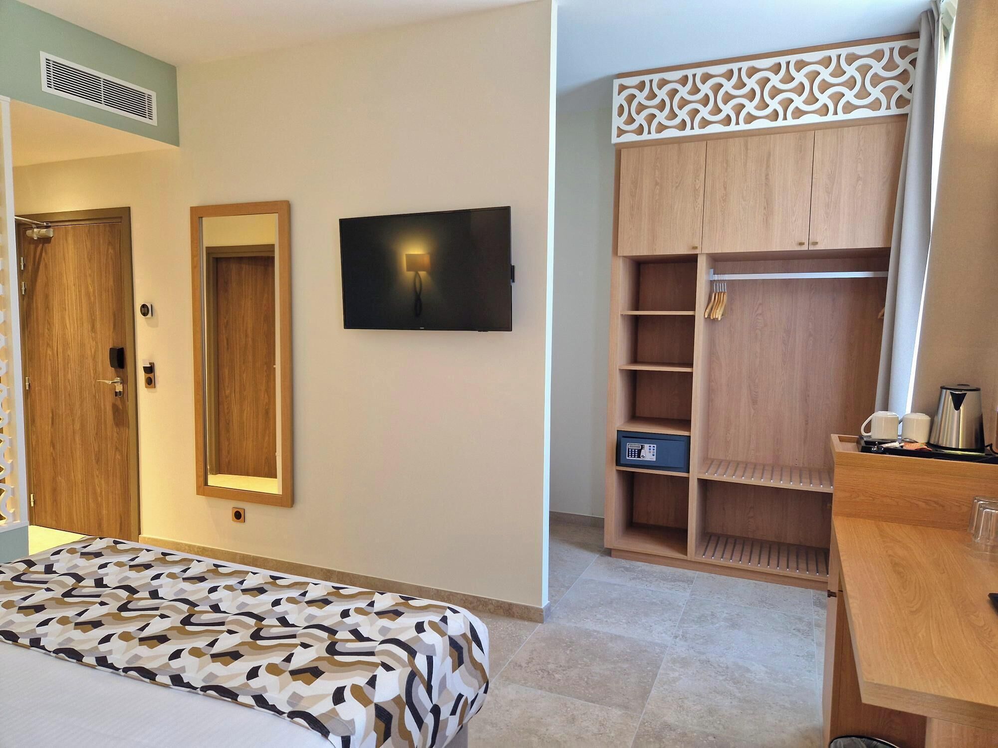 Quarto de hotel com cama, televisão, guarda-roupa e escrivaninha com chaleira e chávenas.