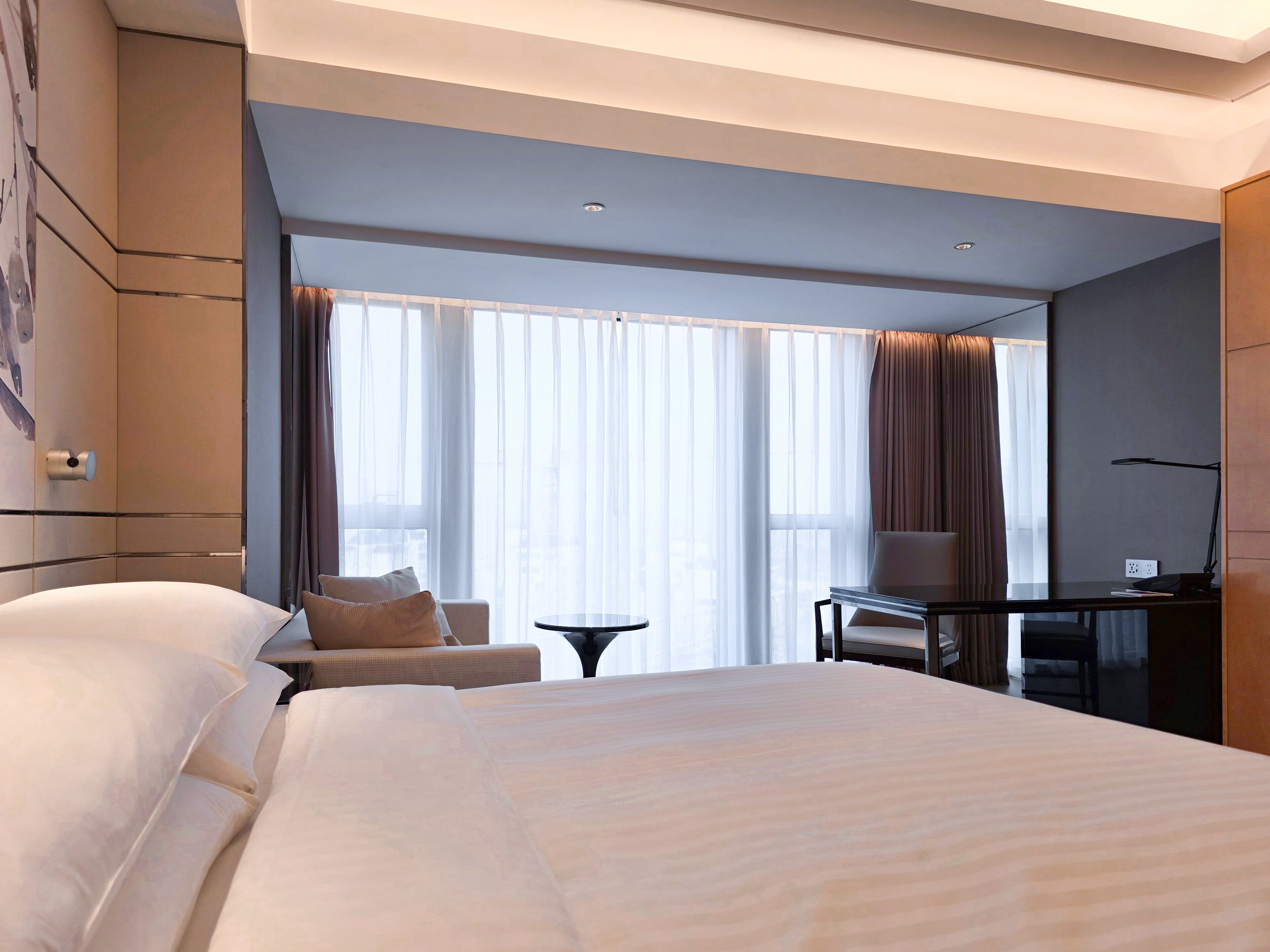 Quarto de hotel com uma cama grande, uma escrivaninha, uma cadeira e vista para a cidade através de amplas janelas.