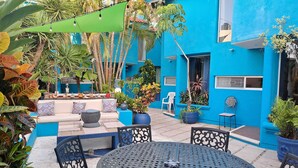 Um edifício azul com mesas e cadeiras ao ar livre, plantas em vasos e uma mesa redonda.
