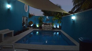 Piscina com parede de pedra e plantas, cercada por um muro azul e uma área sombreada.