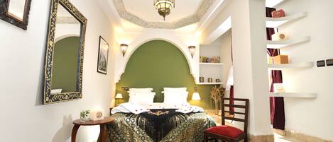 Quarto com arco verde, cama com roupa de cama branca, cadeira de madeira e pequena mesa com abajur.