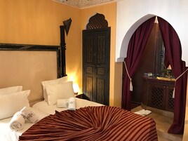 Quarto de hotel com cama, mesa de cabeceira, porta decorativa e banheiro visível através de um arco.