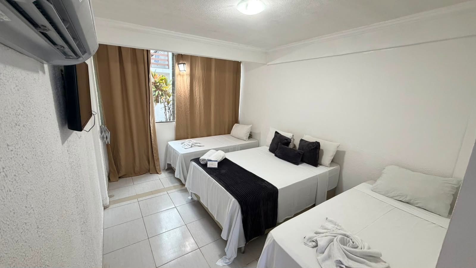 Quarto de hotel com três camas, janela com cortinas, televisão de tela plana e piso de azulejos.