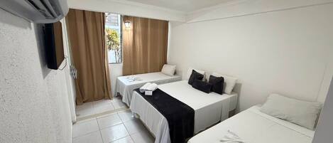Quarto de hotel com três camas, janela com cortinas, televisão de tela plana e piso de azulejos.