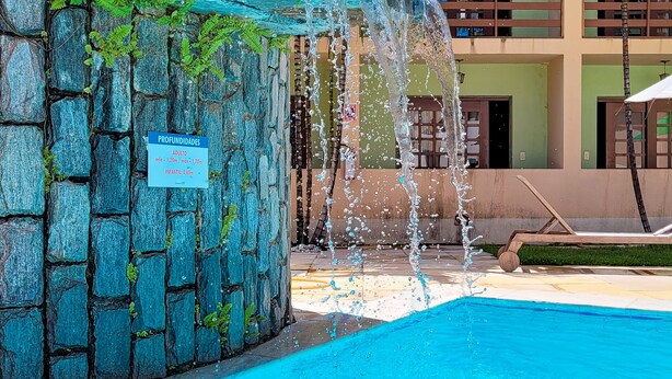 Piscina com cachoeira, placa indicando a profundidade e um edifício com varandas ao fundo.