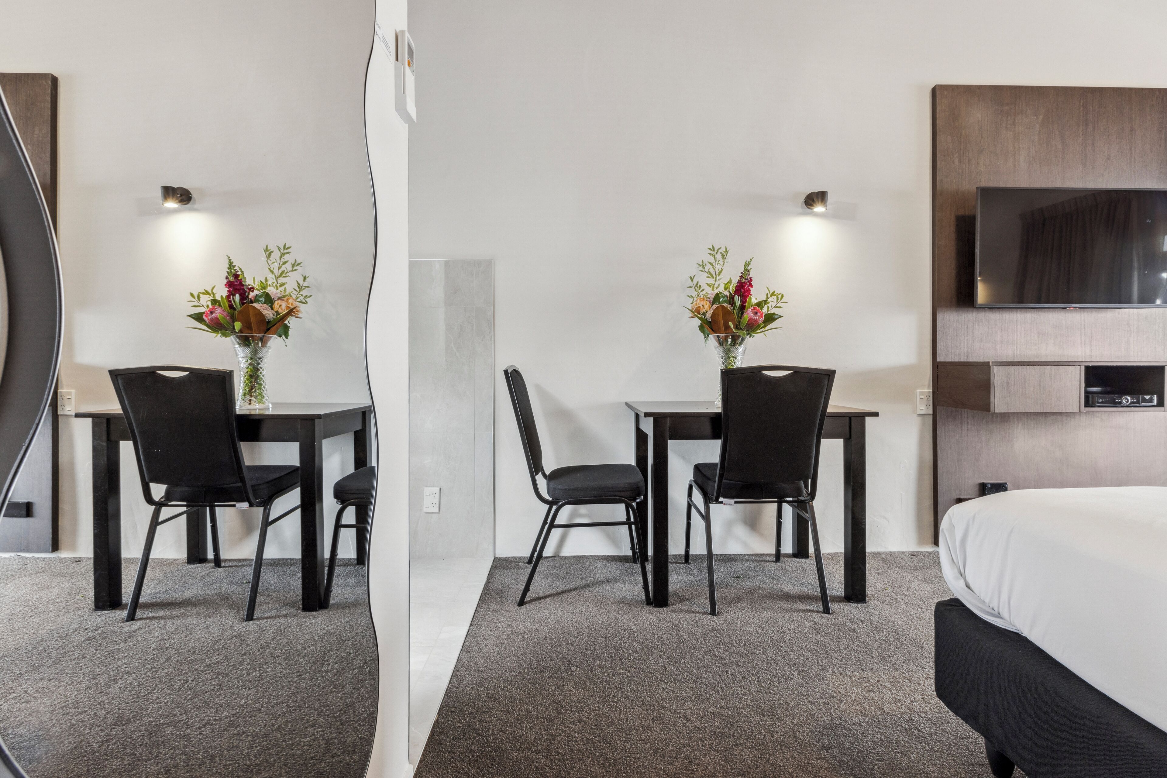 Quarto de hotel com cama, televisão, escrivaninha e duas cadeiras, e dois vasos com flores.
