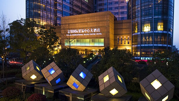 Um hotel moderno com fachada de vidro marcante, entrada central com sinalização iluminada e uma estrutura cúbica distinta em primeiro plano.