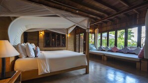 Quarto com cama, mesa de cabeceira, banco com almofadas e grandes janelas com vista para o verde.