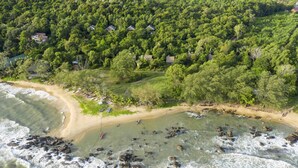 Vista aérea de uma área costeira com praia, costa rochosa e algumas casas rodeadas por floresta densa.