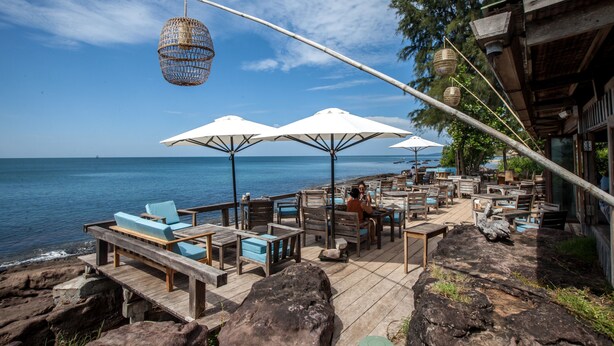 Um deck de madeira com área de estar externa, guarda-sóis brancos e vista para o mar.