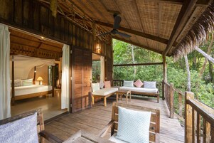 Um deck de madeira com cama, sofá e cadeiras, rodeado por vegetação exuberante.