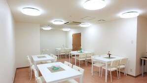 Um quarto amplo com várias mesas e cadeiras brancas, luminárias embutidas no teto e uma porta ao fundo.