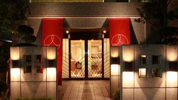Entrada de hotel com caminho de azulejos que leva a um conjunto de portas duplas, ladeada por pilares iluminados e um toldo vermelho.