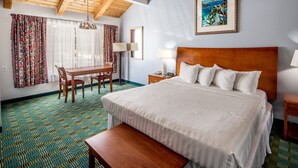 Quarto de hotel com uma cama grande, cabeceira de madeira, uma mesa de jantar com cadeiras, um banco e uma janela ampla com cortinas.