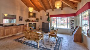 Uma sala de estar ampla com vigas de madeira no teto, lareira, televisão, móveis de vime e uma grande janela com vista para o exterior.