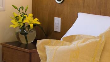 Uma cama com almofadas estampadas em amarelo, uma mesa de cabeceira com um vaso de flores amarelas e uma luminária fixada na parede.
