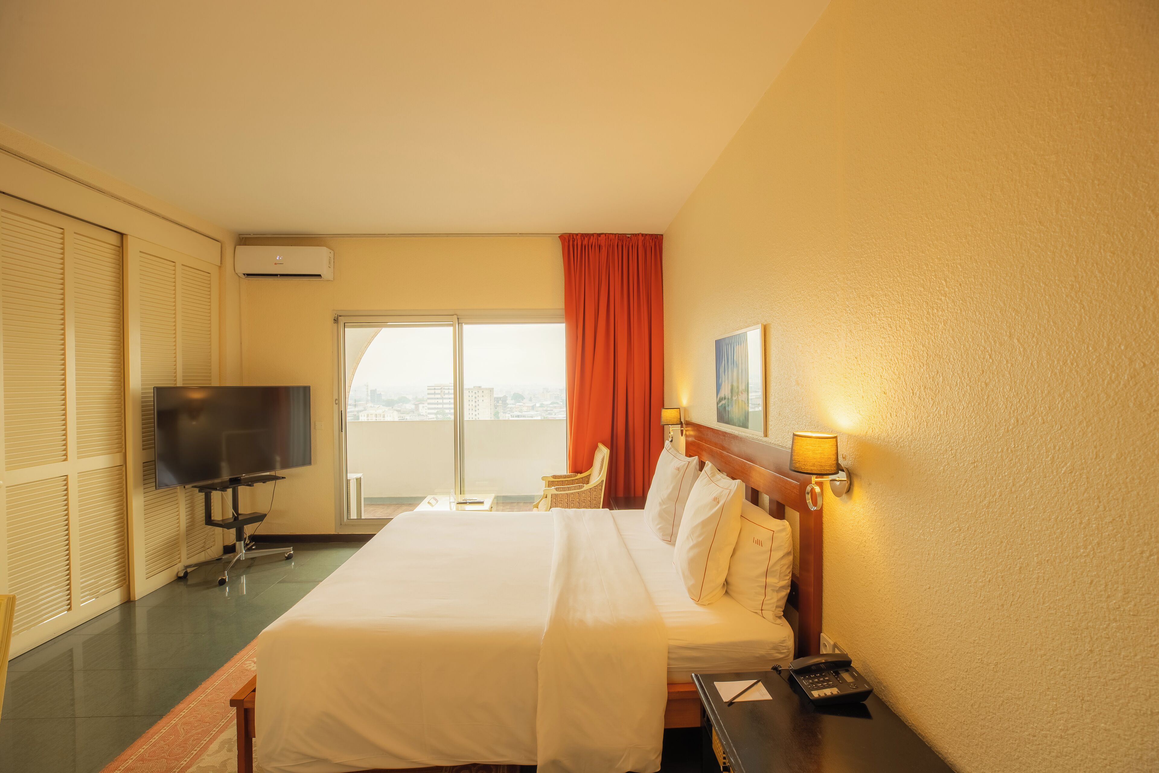 Quarto de hotel com uma cama grande, televisão, escrivaninha com abajur e vista para a cidade pela janela.