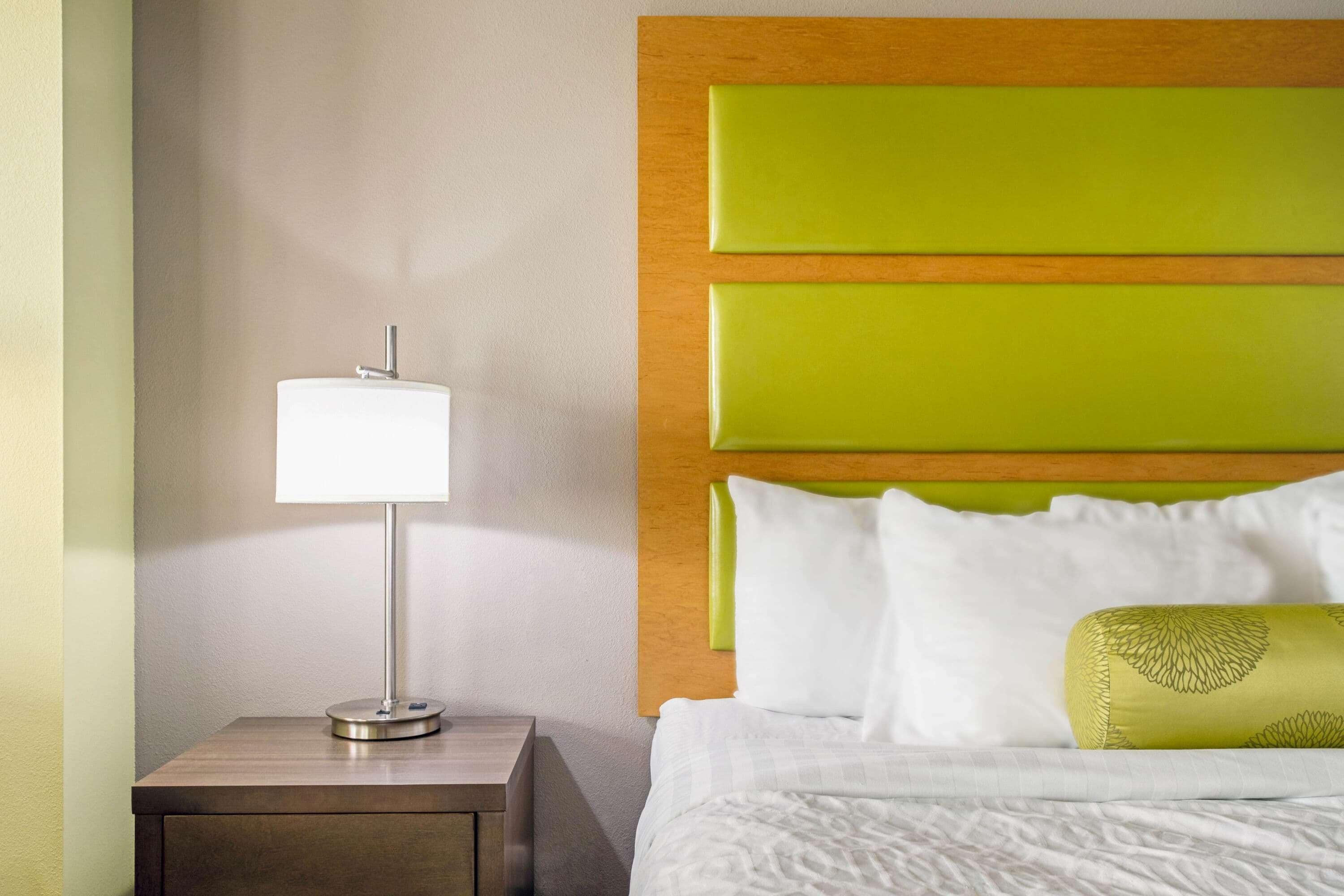 Uma cama bem arrumada com lençóis brancos e um travesseiro verde, uma luminária de cabeceira em um criado-mudo de madeira, e uma cabeceira de madeira com painéis verdes.