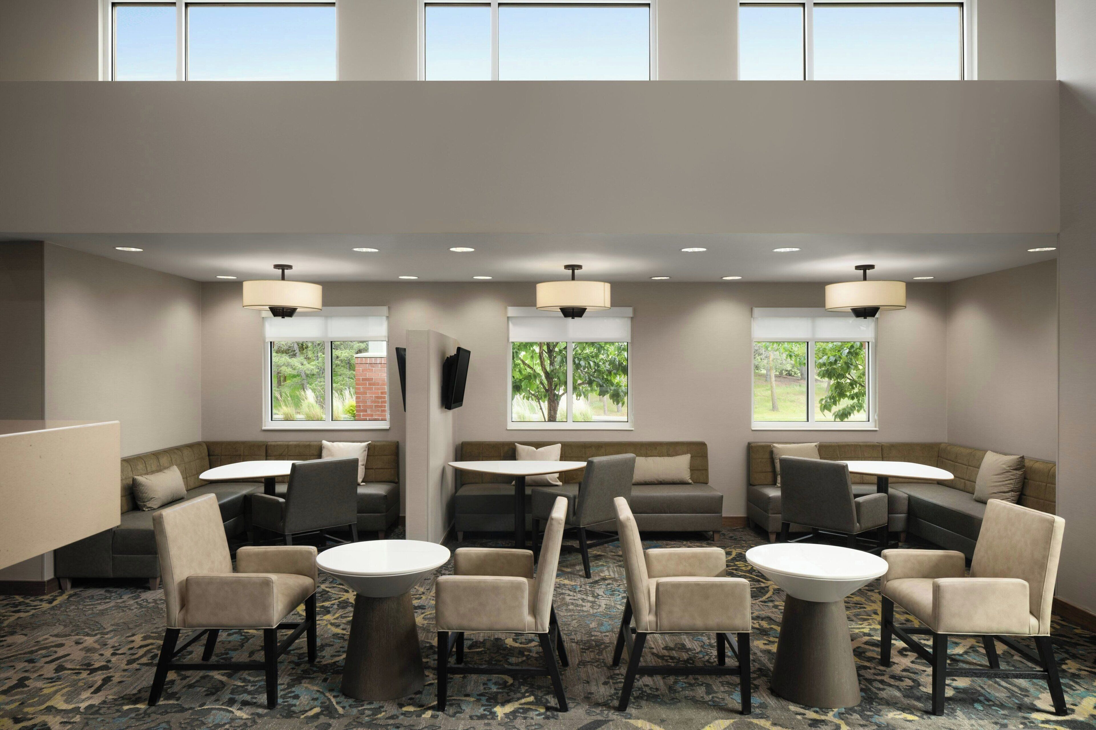 Um hall de hotel com várias áreas de estar, incluindo sofás e cadeiras, dispostas ao redor de mesas. Grandes janelas proporcionam iluminação natural e vista para o verde lá fora.