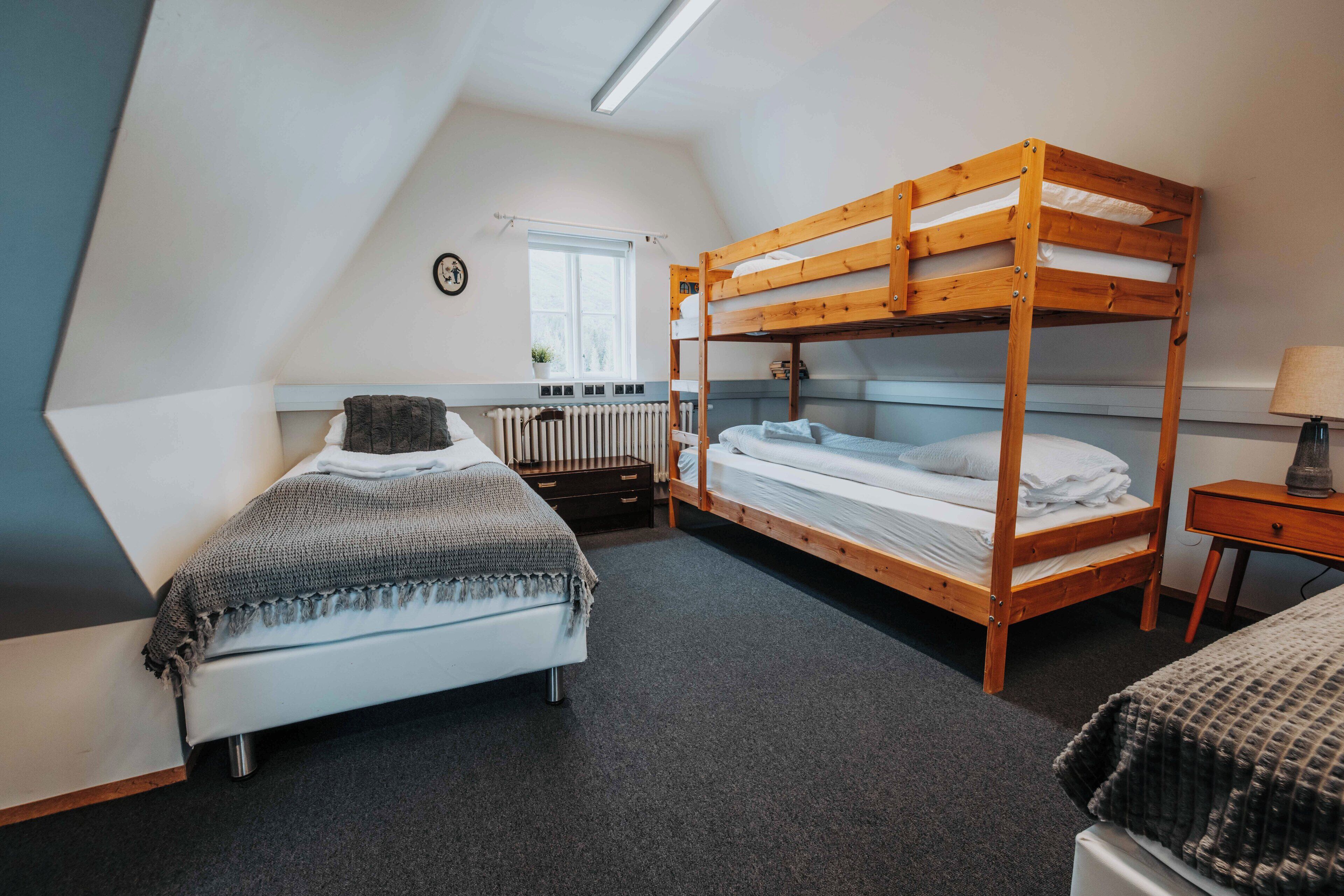 Um quarto de dormitório com uma cama beliche, uma cama de solteiro, um radiador, uma janela e uma escrivaninha com abajur.