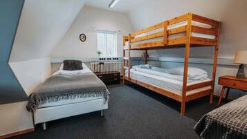 Um quarto de dormitório com uma cama beliche, uma cama de solteiro, um radiador, uma janela e uma escrivaninha com abajur.