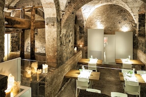 Um restaurante com paredes de pedra e vigas de madeira, com mesas postas com toalhas brancas e velas.